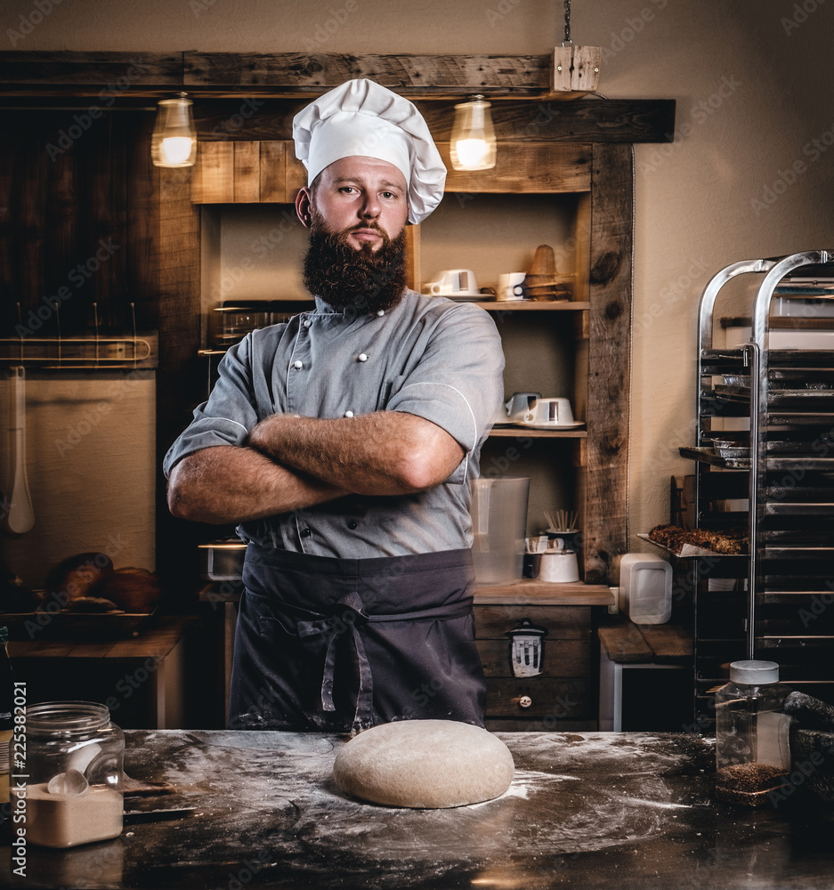 Professional baker in cook uniform posing with crossed arms near table ...