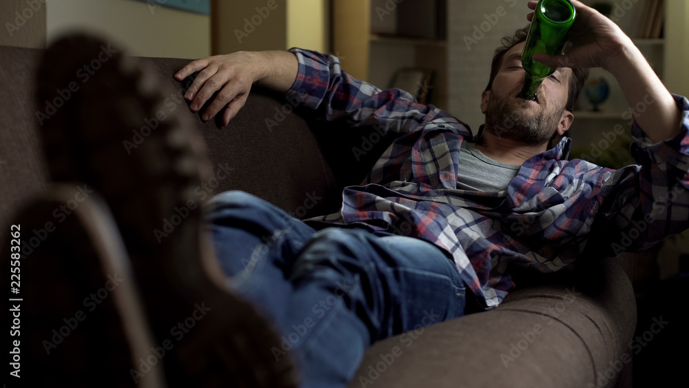 Wasted guy lying on sofa, drinking up beer from bottle, dozing off ...