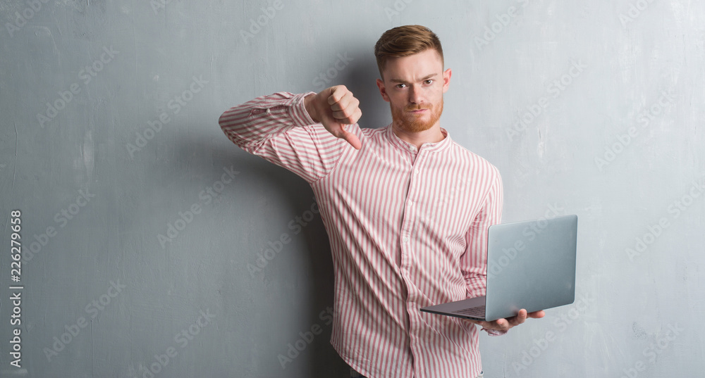 Young redhead man over grey grunge wall holding and using computer ...