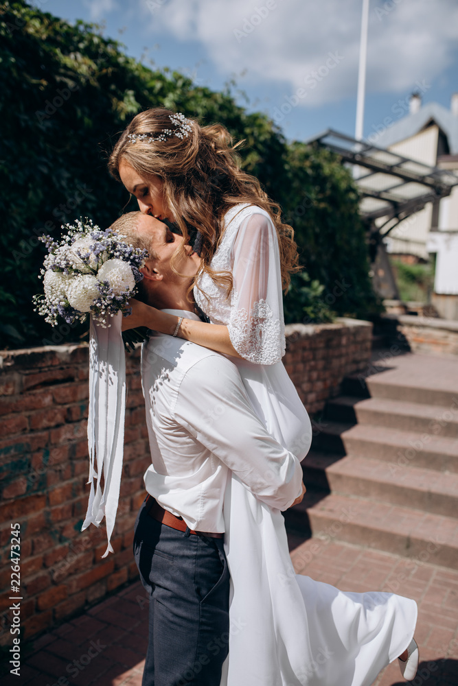 Groom holds bride on his arms tender standing somewhere in an old town ...