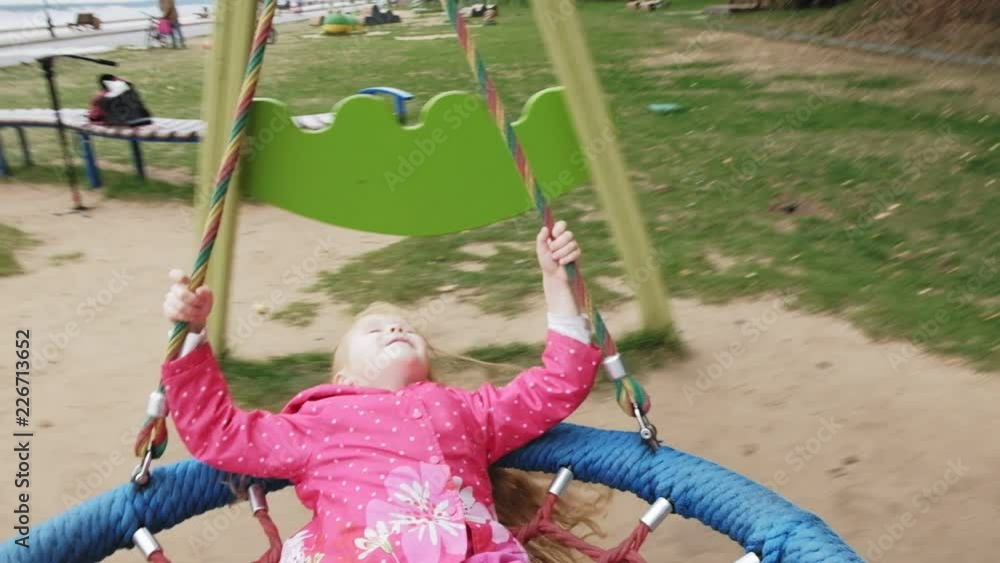A little girl in a pink dress swings on a round swing in the playground ...