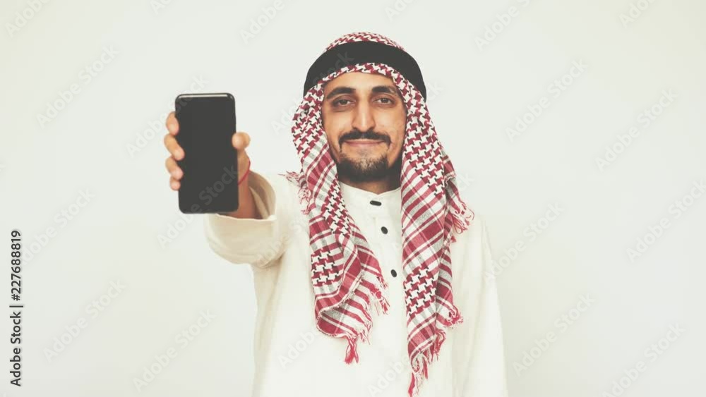 Smiling Arab in national dress shows smartphone and smiles widely. A ...