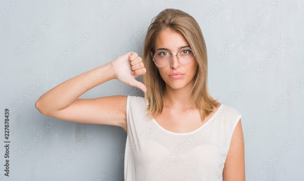 Beautiful young woman standing over grunge grey wall wearing glasses ...
