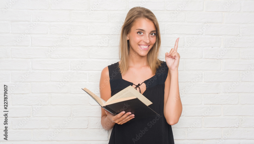 Beautiful young woman over white brick wall reading a book surprised ...