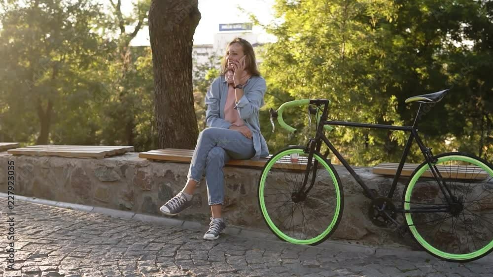 Fair haired girl sitting on the bench or parapet in the city park with ...