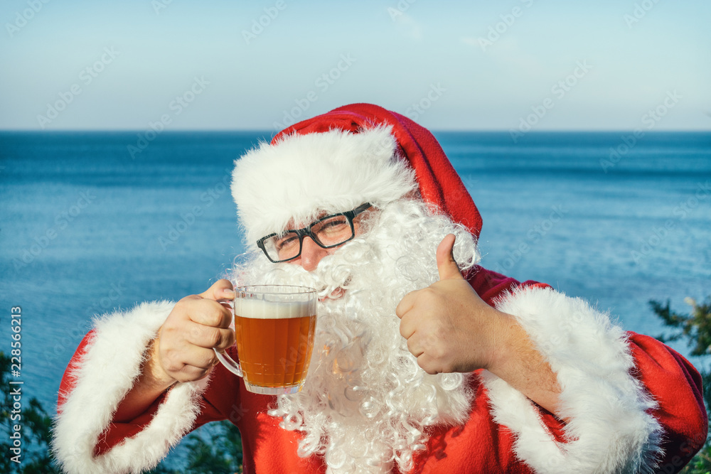 Fat man in glasses dressed as Santa drinking beer on the ocean. Funny ...