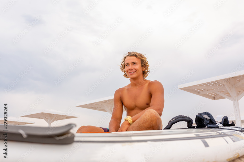 Handsome man sits on the beach with white blank surfing board wait for ...