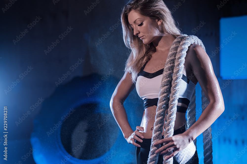 Strong beautiful woman with a battle rope on her shoulder in the gym ...