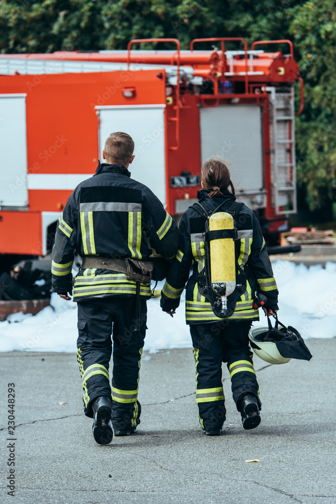 back view of firefighters in fireproof uniform walking on street with ...