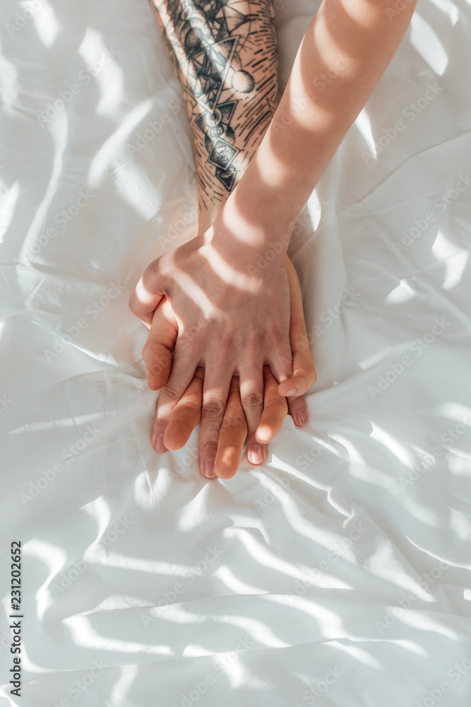 Foto Stock partial view of loving couple holding hands while lying on white bed sheet | Adobe Stock