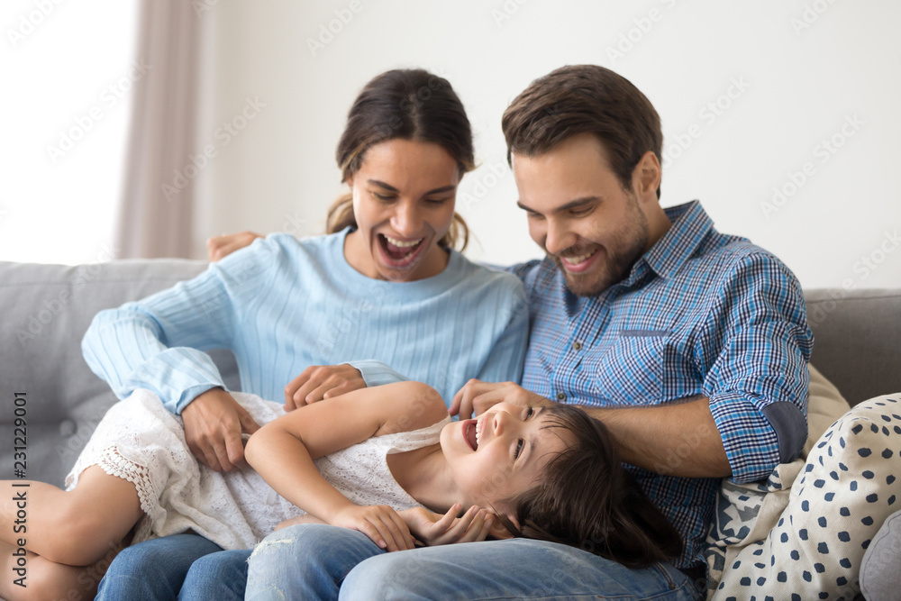 Little funny daughter lying on parents lap while mother father tickling ...