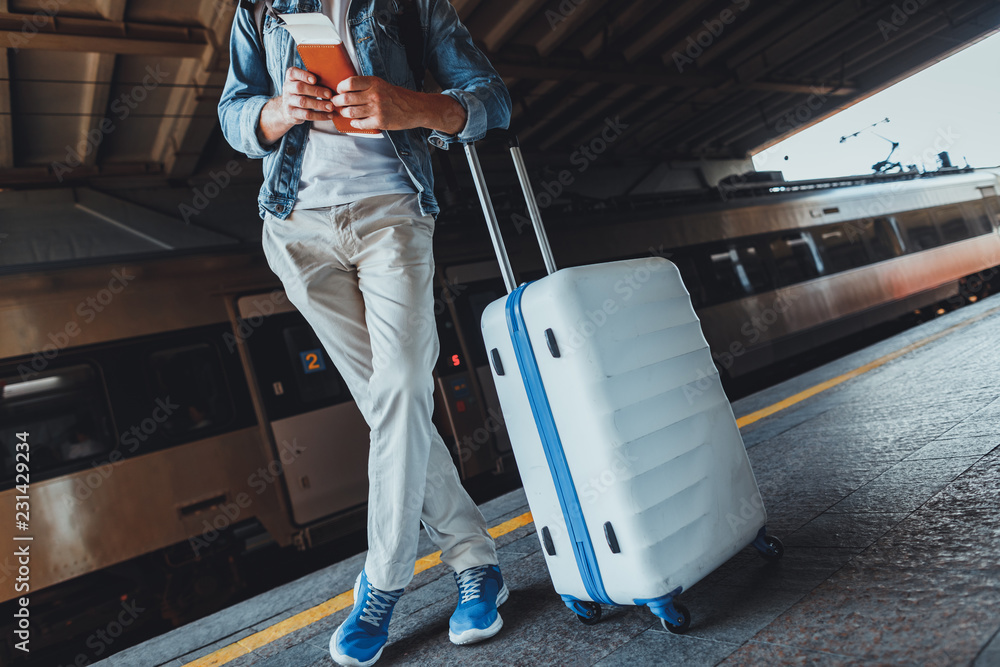 Low angle of a young man standing with his suitcase on the platform of ...