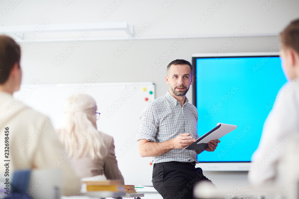 Confident teacher with document looking at group of students while ...