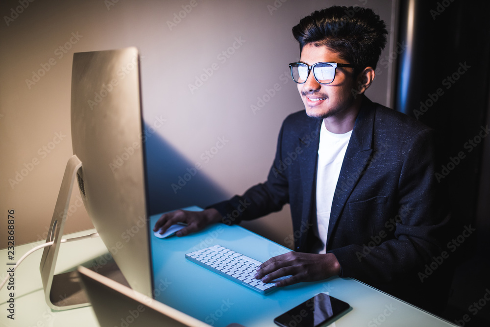 Indian young man sitting and using blank screen computer in dark room ...