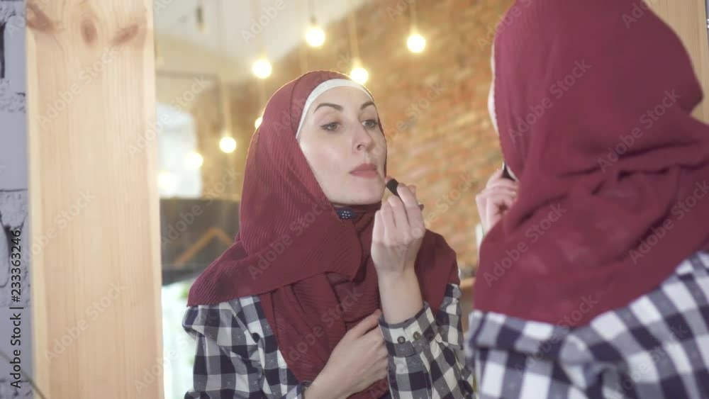 Young muslim woman in a hijab in front of a mirror paints her lips ...