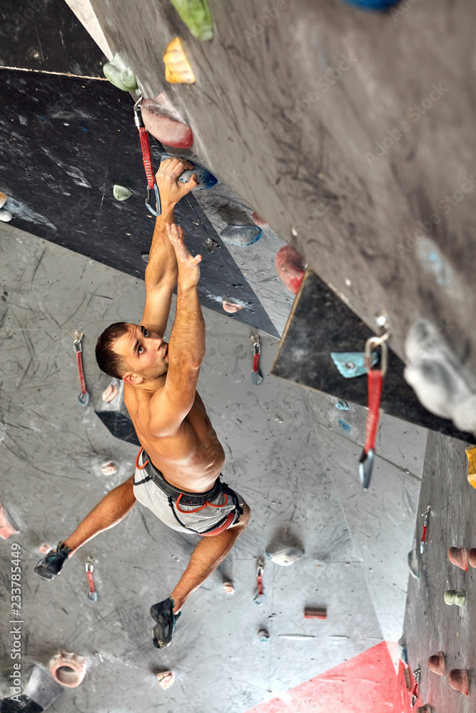 Muscular shitrless man climbing wall with belay device at an indoor ...