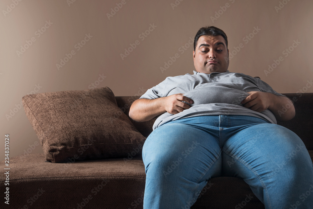 Obese man sitting on sofa holding his belly fat with both hands and ...