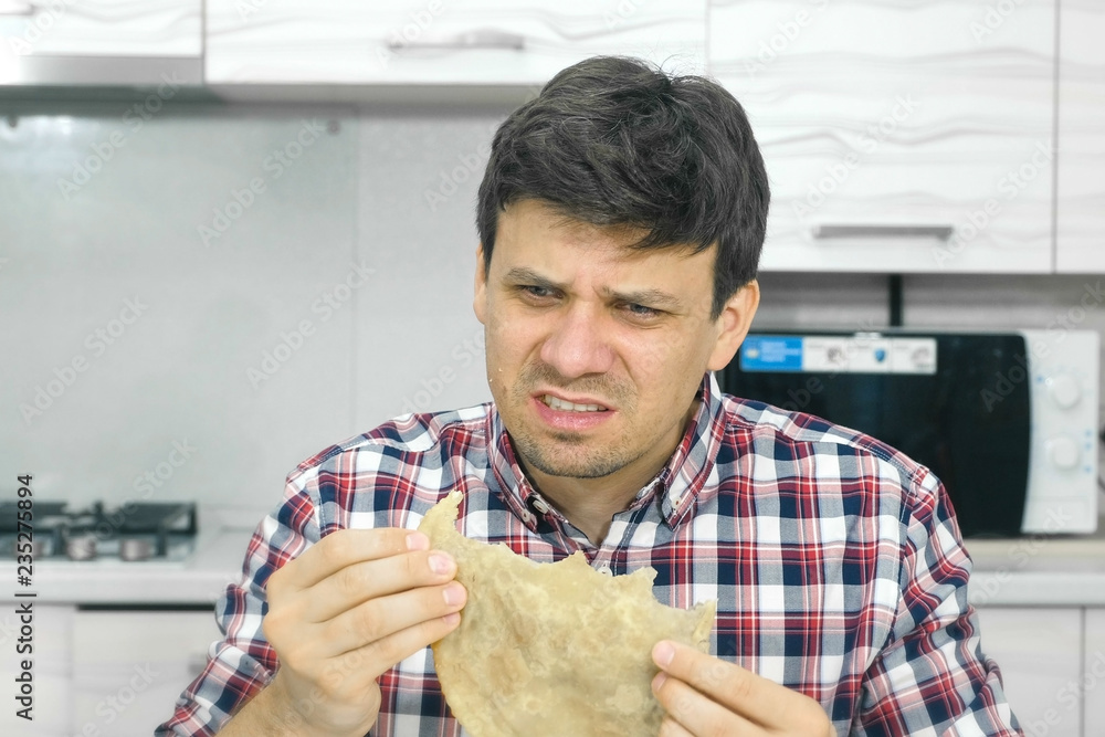 Young man in a plaid shirt eats a big cheburek with disgust and spits ...