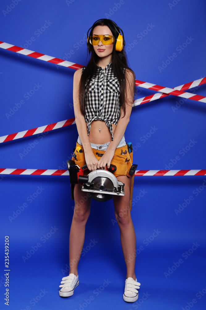 Sexy female construction worker in yellow glasses holding a circular saw on a blue background ...