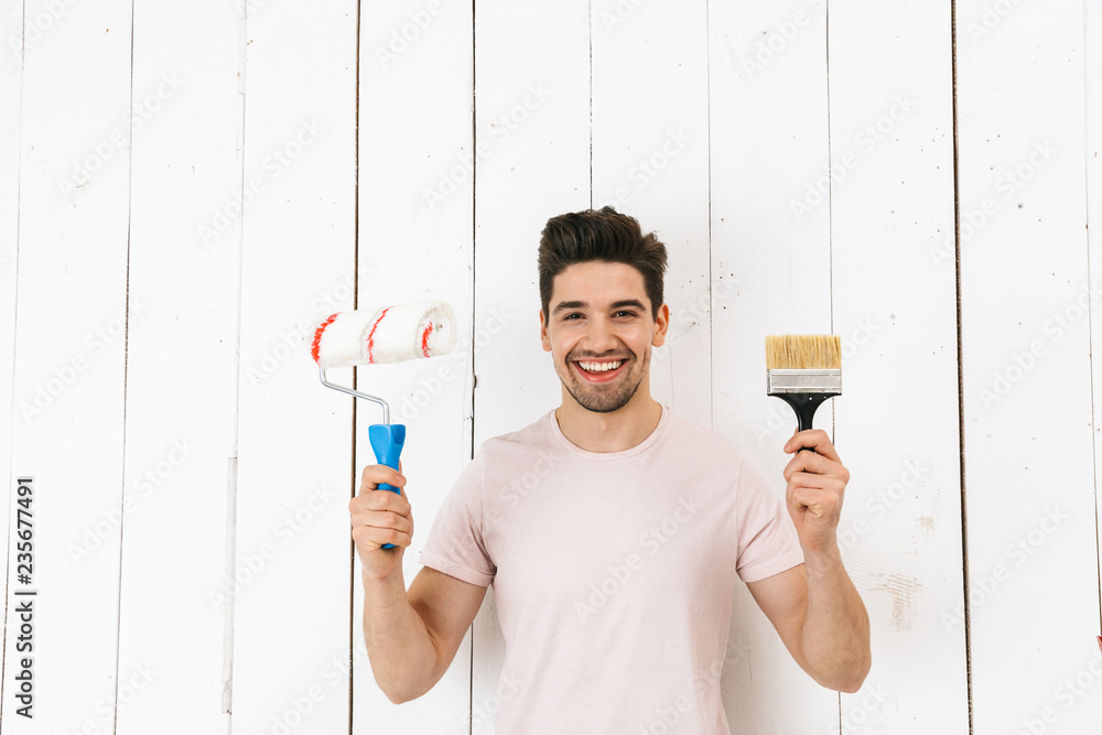 Portrait of joyful man 20s holding paint roller and brush, while ...