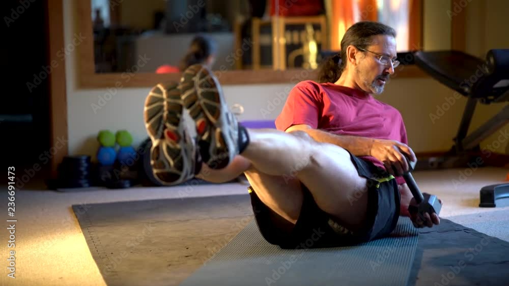 Camera moves right as a mature man uses a barbell while doing russian ...