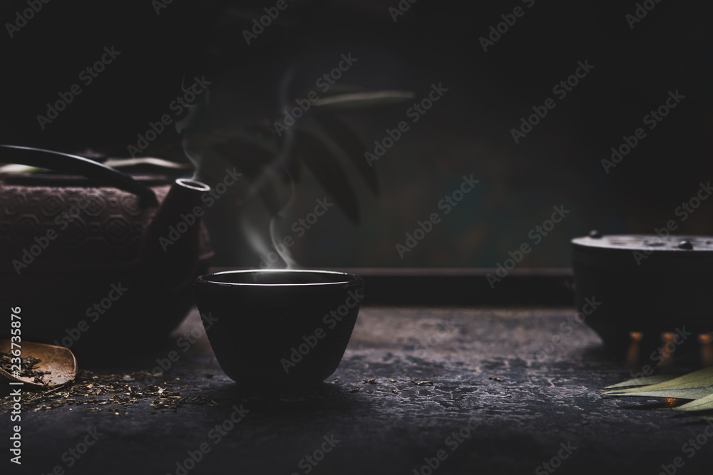 Dark tea background with black iron asian teapot and mug of hot tea ...
