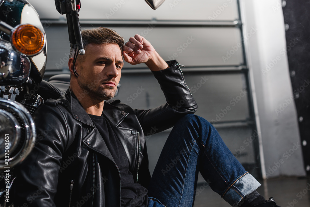 handsome biker looking at camera and leaning on motorbike in garage ...