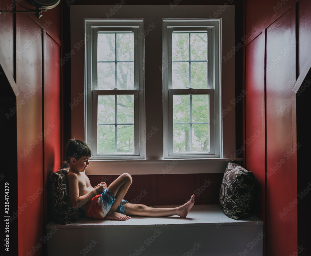 Side view of shirtless cute boy sitting on alcove window seat at home ...