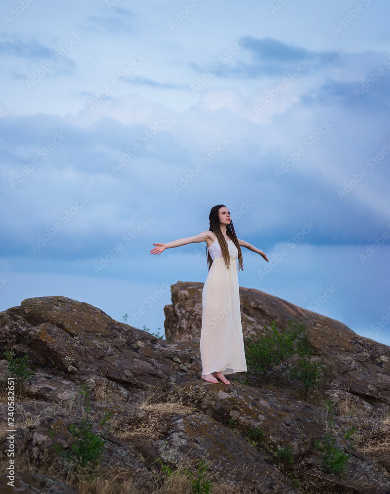 A beautiful girl in a white dress with dreadlocks is standing on a cliff with her arms ...