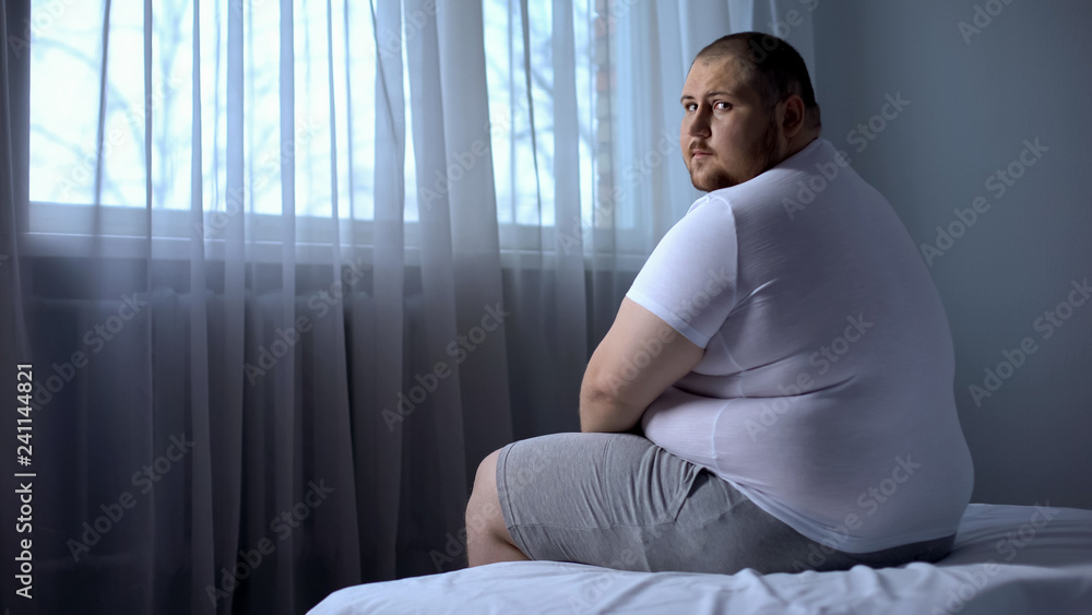 Sad fat man sitting on bed at home, looking at camera, depression ...