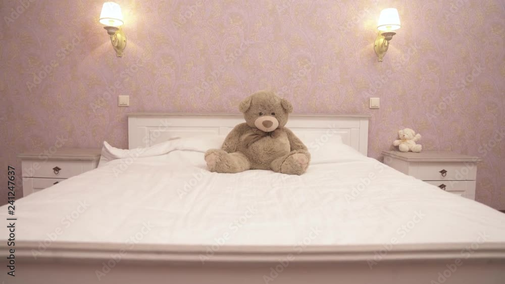 A big teddy bear is sitting on the bed covered with white blanket in the bedroom. The camera ...