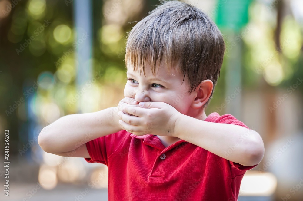 Funny Caucasian child closing his mouth to hide a smile in the park ...
