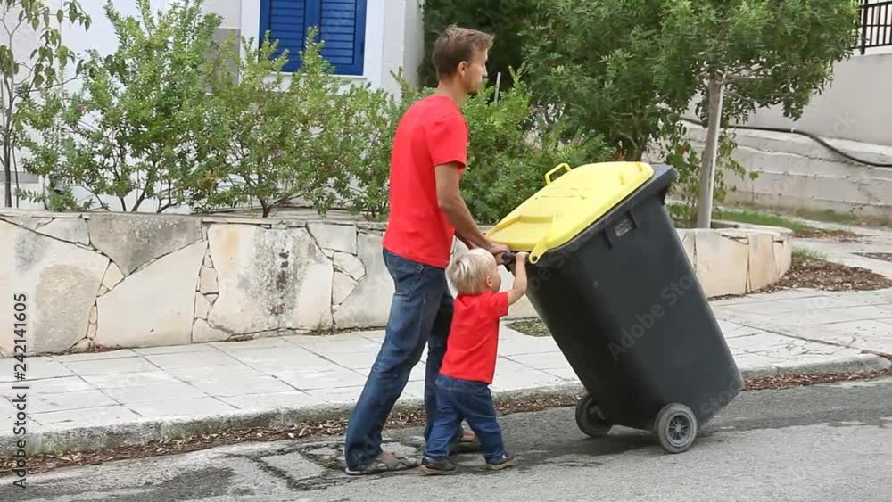 Father and son together push big trash can on wheels on the street ...