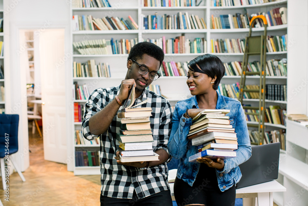 Young smiling african boy and girl holding books standing together on the background of shelves ...