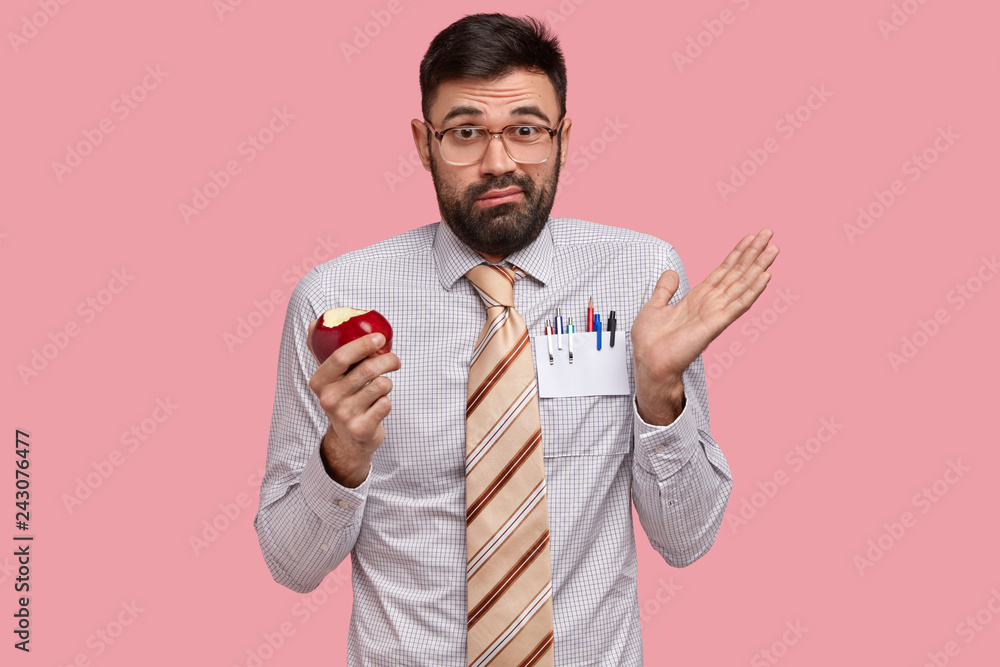 Clueless discontent bearded man spreads hand, holds red apple, wears ...