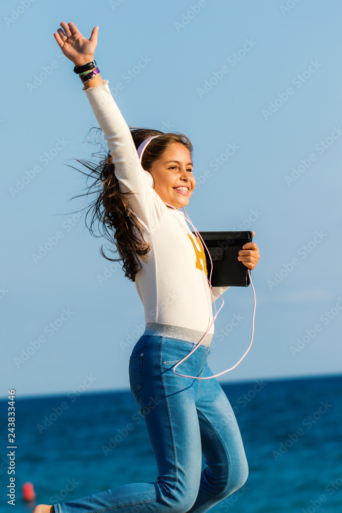 Happy young tween girl jumping with headphones and tablet on beach ...