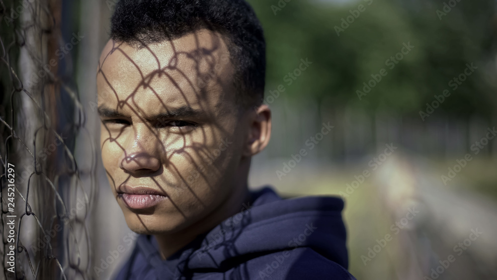 Poor afro-american teenager standing by metal fence, life difficulties ...