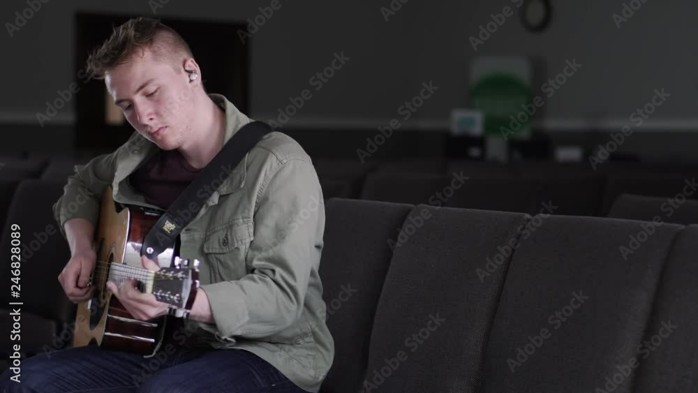 A young man playing an acoustic guitar, alone in a room full of seats ...