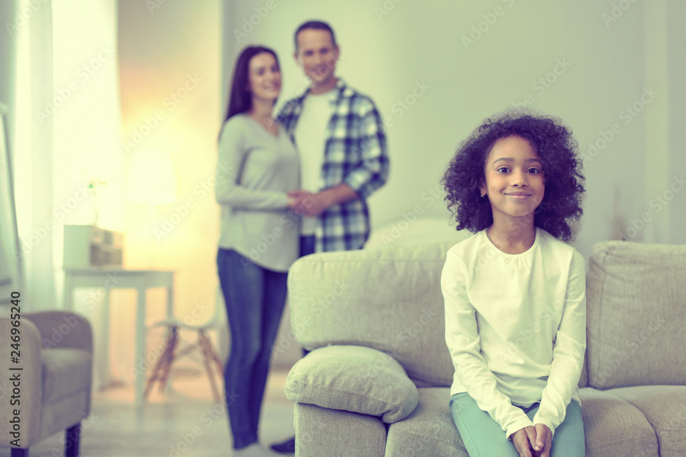 Little girl sitting on the couch with her parents standing behind her ...