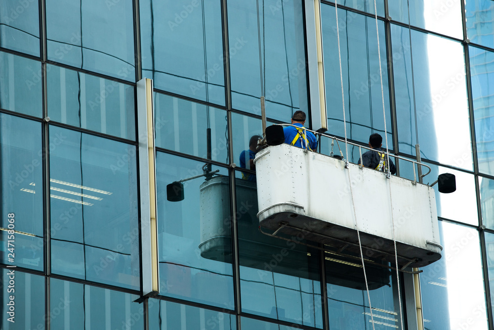 Workers crane cradle clean windows glass of high building under sun day ...