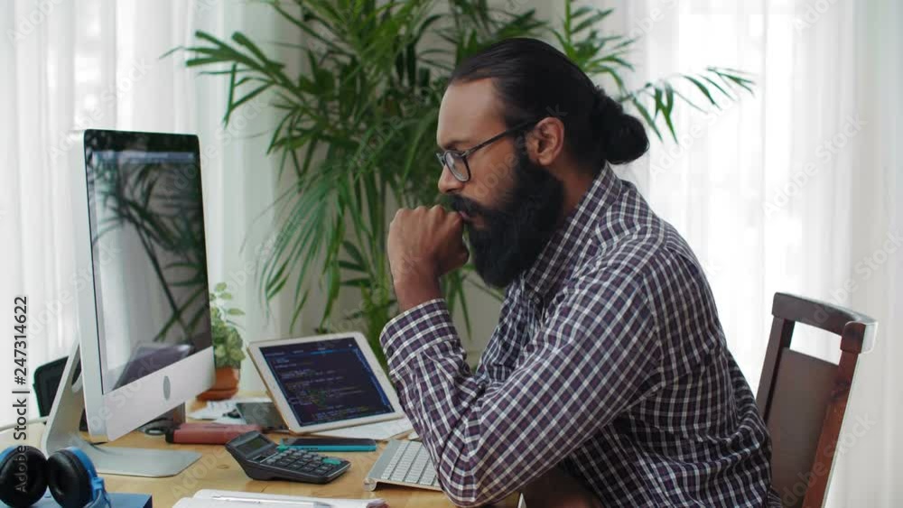 Medium shot of Indian man sitting at worktable, typing software code ...