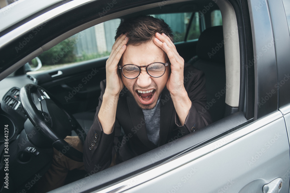 Shocked young guy is sitting at his car. His hands are on his head, his ...