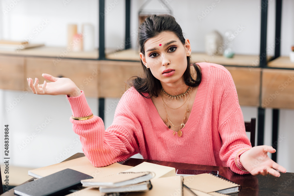 sad indian student with bindi studying with notebooks and showing shrug ...