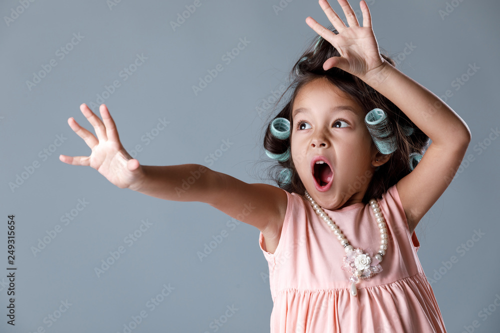 scared little child girl in pink dress and hair curlers posing on gray ...