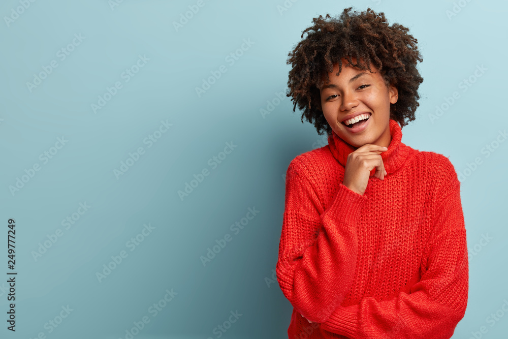 Carefree young woman with dark skin, keeps hand under chin, laughs ...