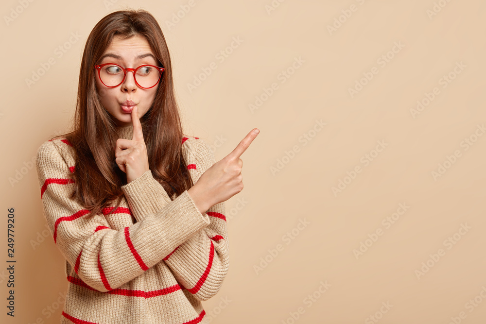 Puzzled lovely woman with curious facial expression, points aside with fore finger, wears ...