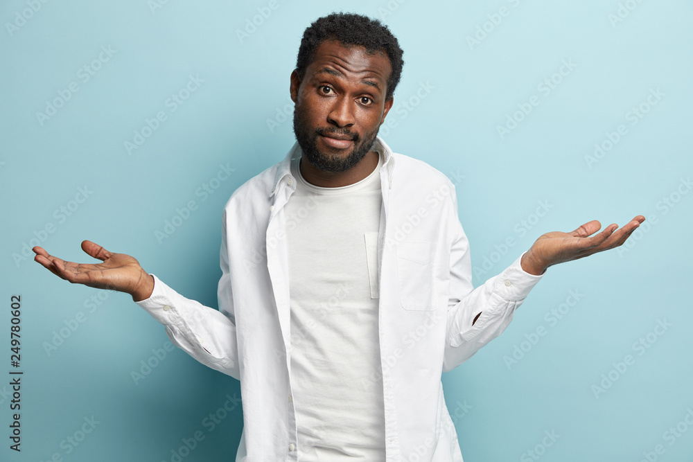 Attractive black ethnic man spreads hands and looks with indecisive ...