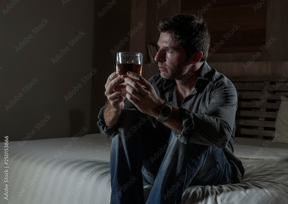 young messy and wasted drunk man drinking whiskey glass at home sitting on bed hammered and ...