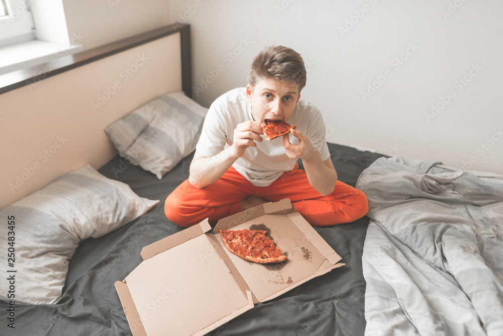 Happy, attractive young man sits home in a cozy room on a pizza box bed ...