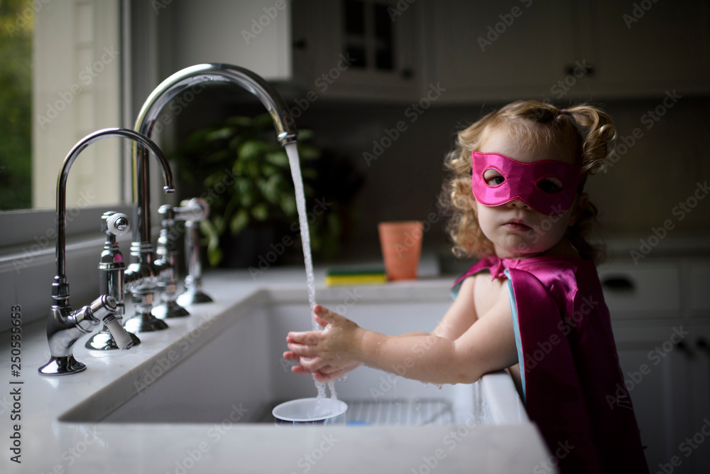 Portrait of girl in superhero costume washing hands in kitchen sink ...